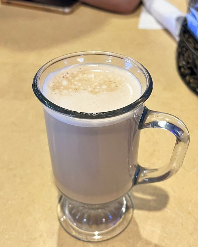 The cappuccino arrives in a glass mug that showcases its perfect layers – strong espresso foundation, velvety milk, and cloud-like foam cap.