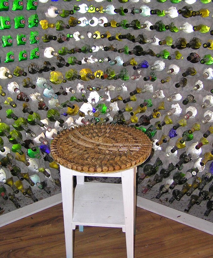 A simple chair with a woven seat sits against a backdrop of bottles arranged in mesmerizing patterns, offering a moment of contemplation amid the glass mosaic.