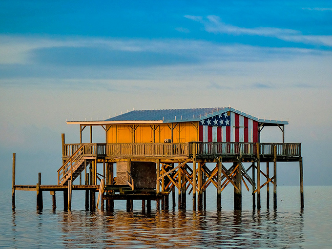 Sunset gilds this American-flagged stilt house in golden light, transforming humble wood into a magical floating fortress.