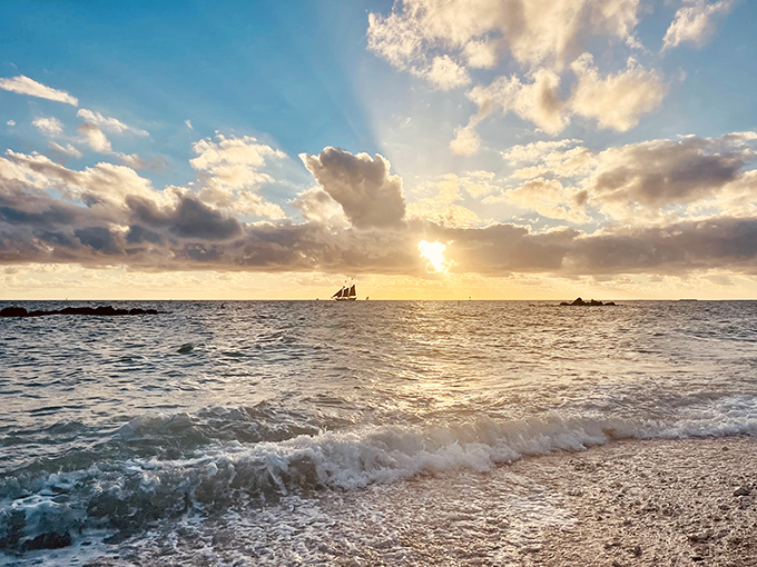 As the day melts into evening, the beach transforms into nature's theater, with sailboats performing against a painted sky.