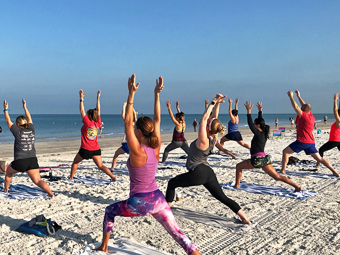 A serene gathering of beach yogis in warrior poses, their silhouettes striking against the calm Gulf waters as morning light bathes everything in golden warmth.