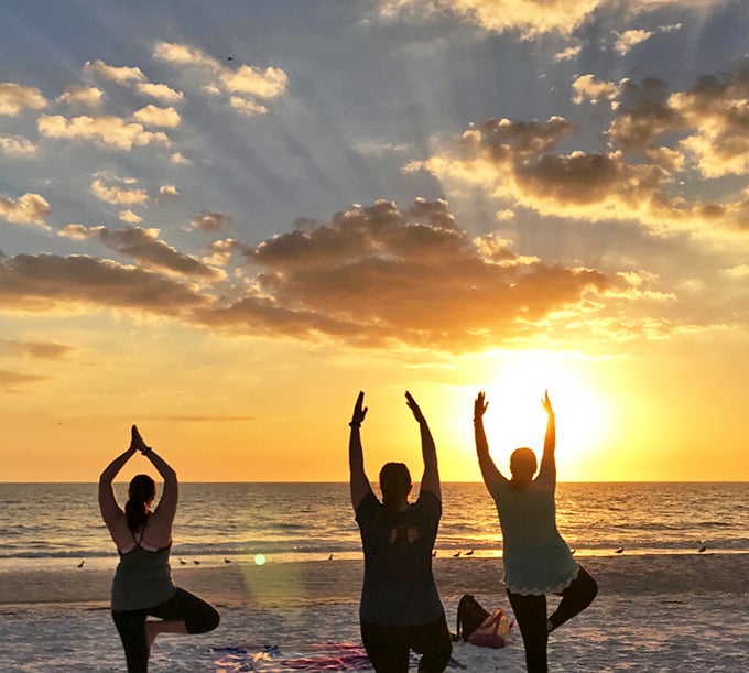 Three silhouettes in tree pose reach skyward as the sun melts into the horizon, turning the Gulf into a mirror of molten gold and the clouds into cotton candy.