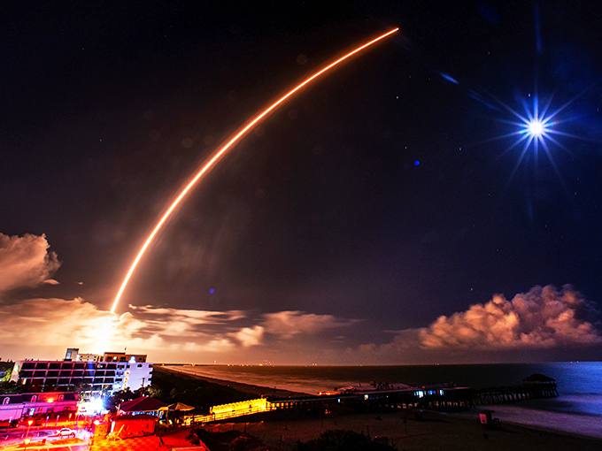 A SpaceX rocket climbs skyward, painting a golden trail against twilight clouds – just another magical evening for lucky Rikki Tiki patrons.