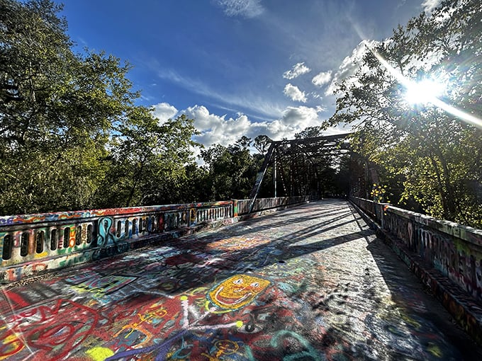 Sunlight filters through the metal framework, casting geometric shadows that dance across the rainbow canvas of accumulated expression.