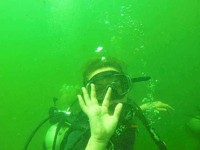 Suspended in the emerald waters, a diver waves to the camera, surrounded by the gentle dance of filtered sunlight.