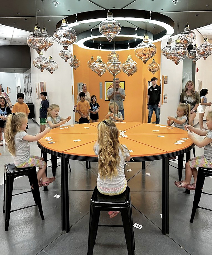 Children gather around the Infinite Reflections Table, where hanging ornaments multiply endlessly in a mesmerizing display.