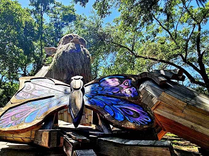 A magnificent wooden butterfly sculpture spreads its vibrant wings against the backdrop of towering trees, nature's artistry meeting human creativity at Folly Farm.