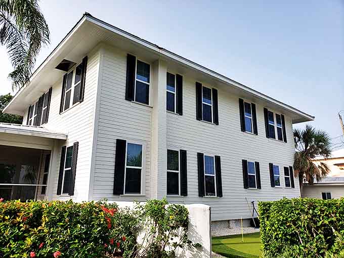 Admire the clean lines of this classic white house with dark shutters, perfectly framed by lush green hedges and flowers.