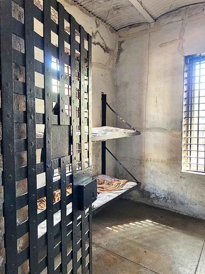 Sunlight streams through barred windows into a sparse cell, illuminating the harsh simplicity of prisoner accommodations that haven't changed in a century.