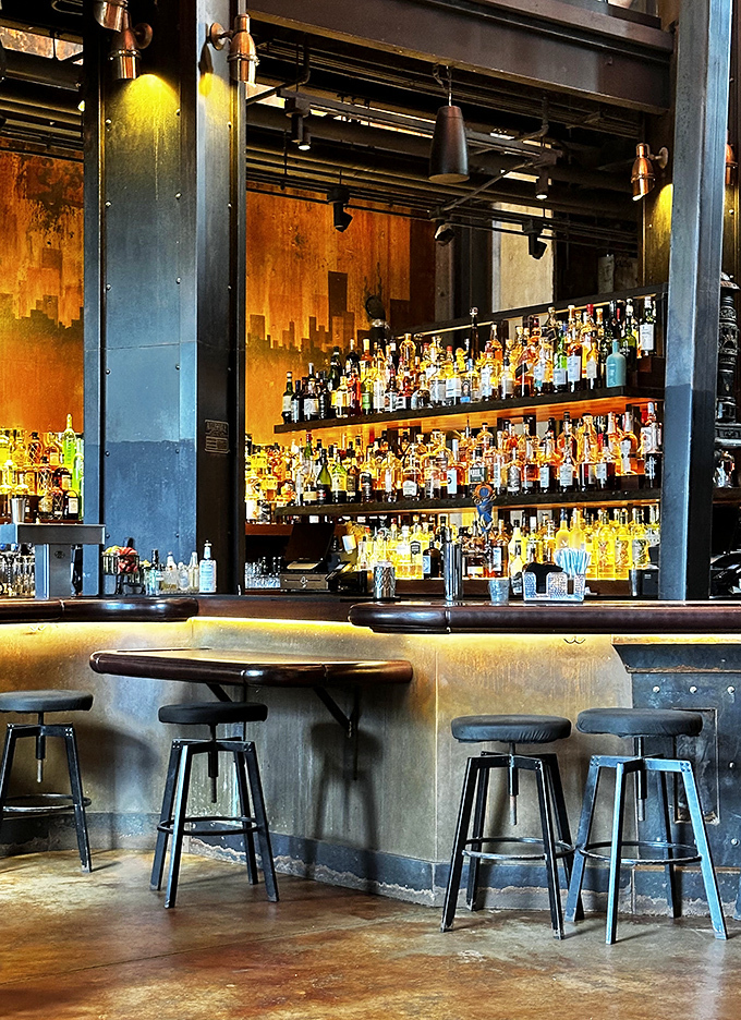 The Edison's bar gleams with amber-lit bottles against industrial metal, where mixologists perform liquid alchemy beneath exposed pipes and vintage gauges.