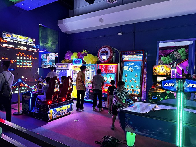 Arcade enthusiasts young and old stand transfixed by flashing screens, their faces illuminated by the glow of digital challenges.