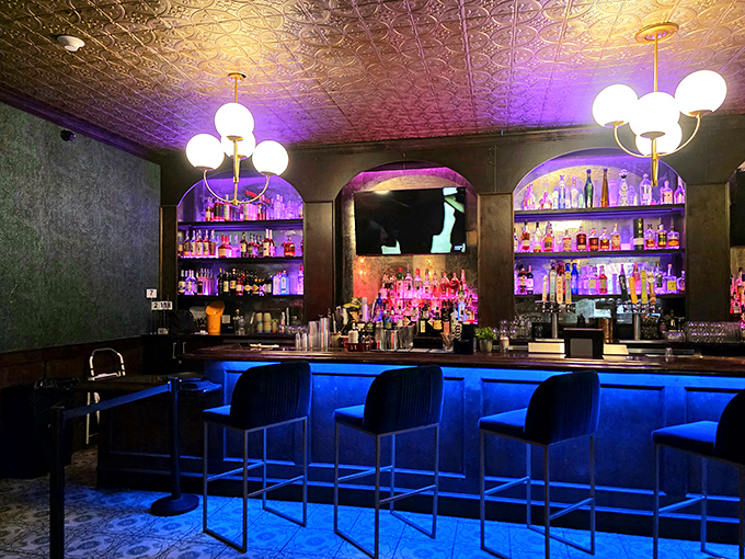 Illuminated in cool blue light, the speakeasy's bar gleams with promise, globe lights casting a warm glow over a sophisticated selection of spirits.