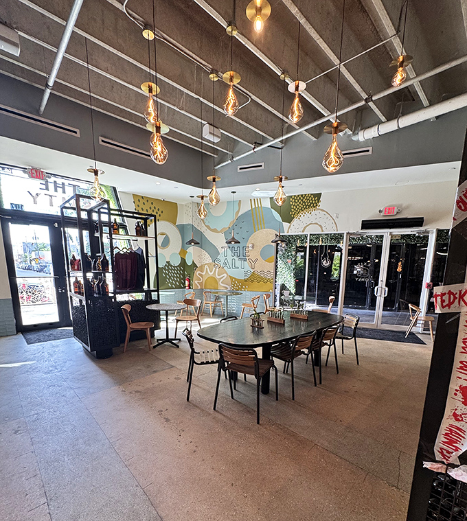 Interior: Edison bulbs dangle like golden jewels above thoughtfully arranged tables where strangers become friends united by their donut devotion.