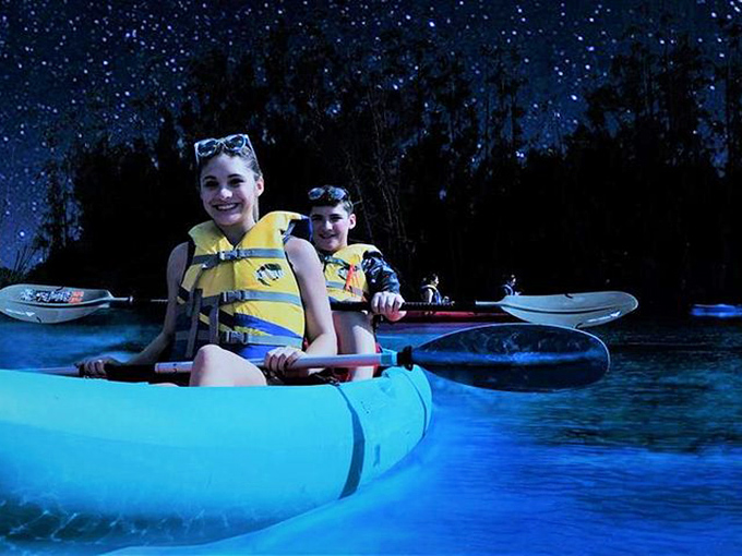 Adventurers glide through liquid starlight, their yellow vests popping against the supernatural blue glow beneath their boats.