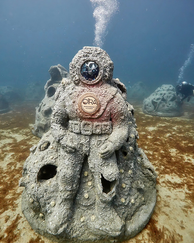 An ORA-branded sculpture stands sentinel on the ocean floor, its purpose both artistic and ecological as marine life begins to claim it.