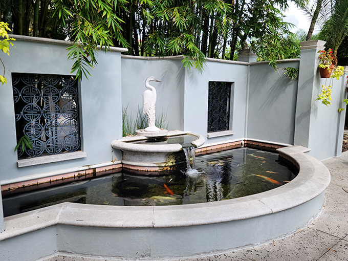 This elegant heron presides over its reflecting pool kingdom, where koi fish dart beneath the surface like living jewels in liquid glass.