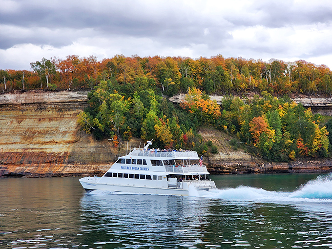 The Pictured Rocks cruise boat looks almost toy-like against the massive autumn-painted cliffs, giving perspective to nature's grand scale.