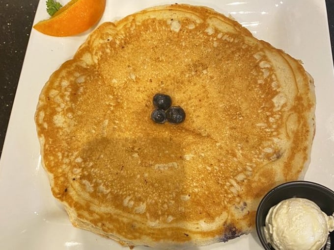 This blueberry pancake is studded with bursts of fruit that pop with each bite, complemented by a side of butter and fresh orange.