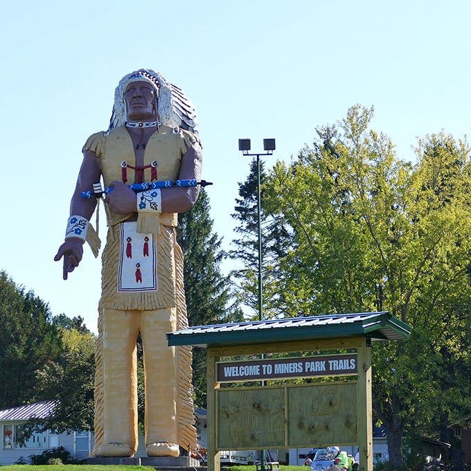 The "Welcome to Miners Park Trails" sign stands dwarfed by its famous neighbor, inviting visitors to explore beyond the statue into the area's natural beauty.