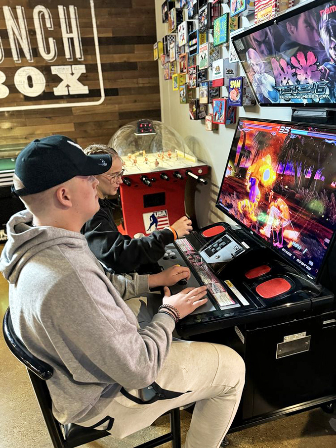 Intense concentration meets explosive on-screen action as gamers dive into a fighting game battle, while the nostalgic wall of gaming memorabilia stands witness to this timeless arcade ritual.