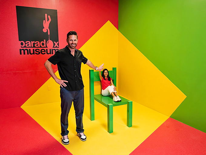 Perspective magic makes a normal-sized chair transform adults into miniature versions of themselves. It's like Alice in Wonderland's growth spurt in reverse, minus the questionable mushrooms.