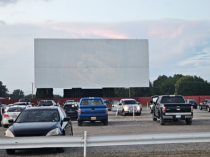 The massive white screen of the Capri Drive-In stands ready against the twilight sky, cars gathering like fireflies at dusk for the evening's cinematic journey.