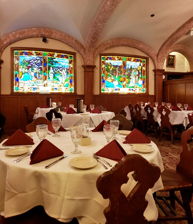 Interior other angle: Stained glass windows cast colorful light across tables set with precision, while wood paneling and heraldic shields transport diners to another era.