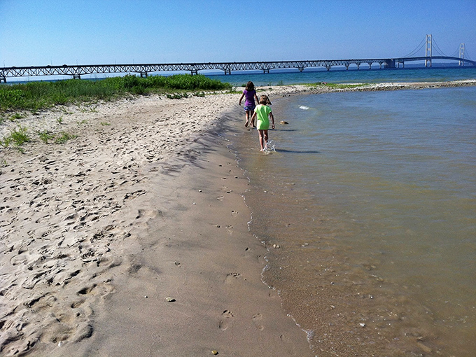 Children explore the shoreline where bridge meets land, their footprints temporary compared to the Mighty Mac's enduring presence.