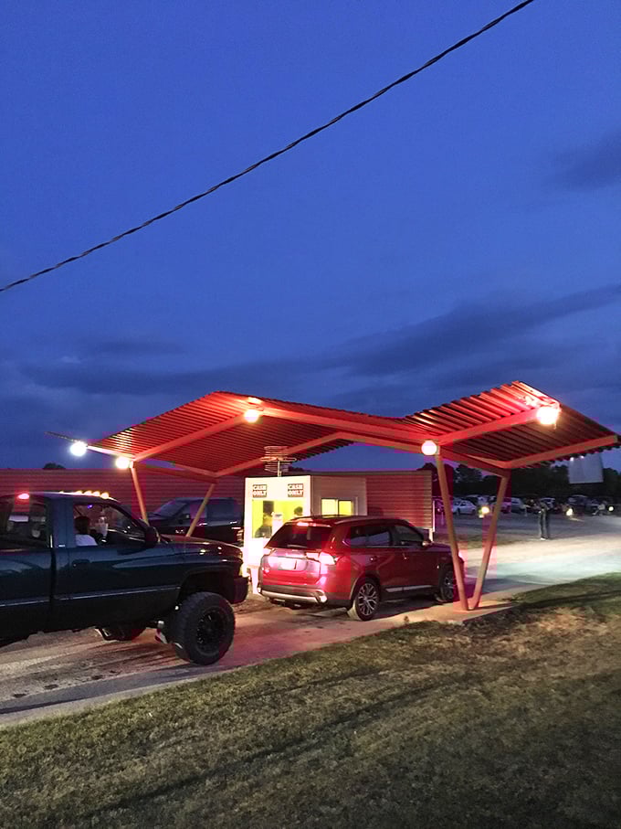 The retro-futuristic ticket booth glows with welcoming red light, a portal transporting visitors from everyday life to drive-in movie magic.