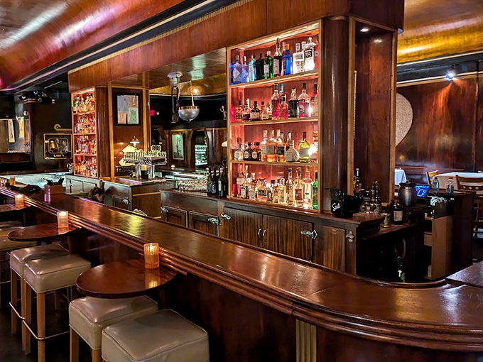 The bar curves like a wooden wave, bottles backlit like precious artifacts waiting to be transformed into liquid magic by skilled hands.