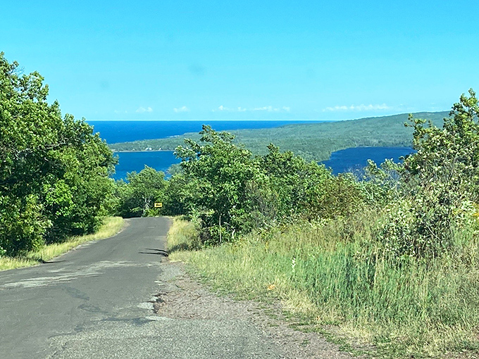 The road curves like a lover's smile, revealing Lake Superior's sapphire waters embracing the distant shoreline in a timeless dance.
