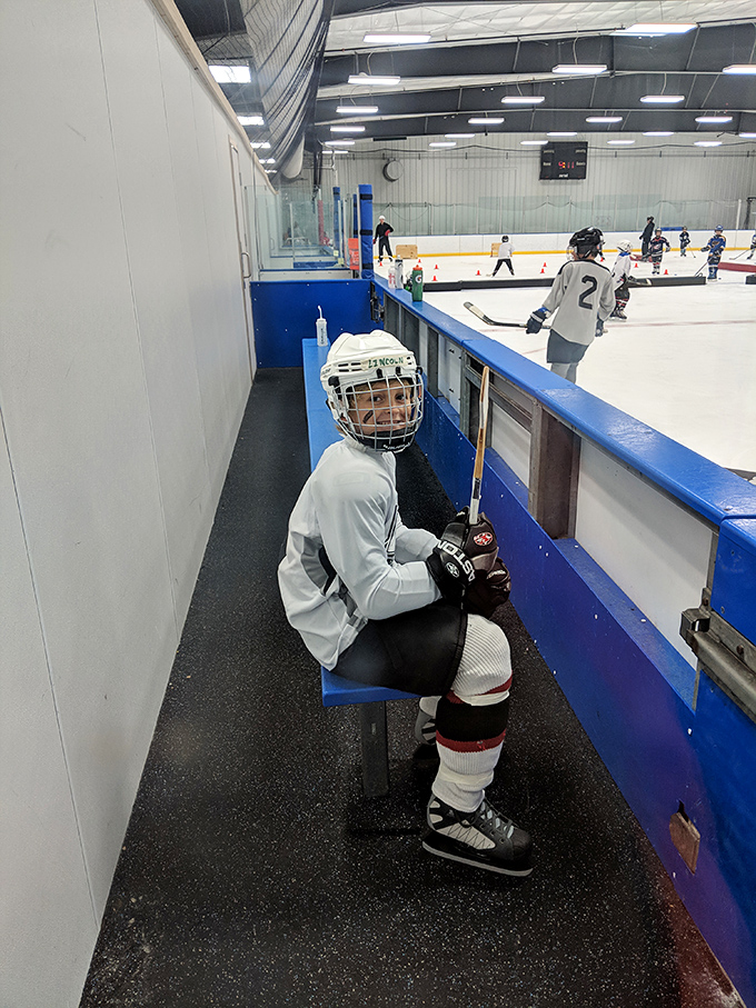 A young hockey player waits for his turn on the ice, equipment ready and anticipation visible even behind the helmet.