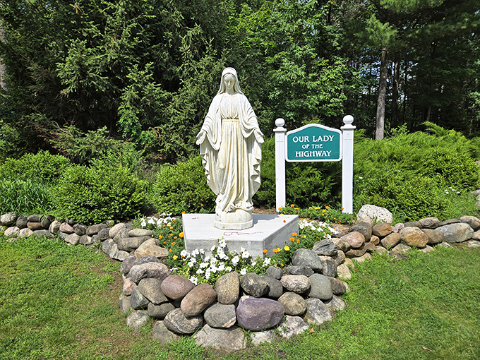 Our Lady of the Highway stands serene among flowers and fieldstones, offering a peaceful roadside blessing to travelers passing by.