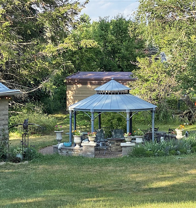 A gazebo nestled among mature trees offers a shaded retreat for afternoon reading or morning coffee amid the garden's natural beauty.