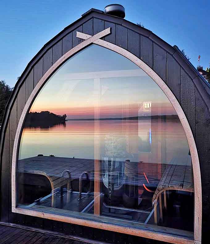 Twilight transforms the sauna into a glowing lantern on water, its arched window reflecting both inner warmth and outer beauty.