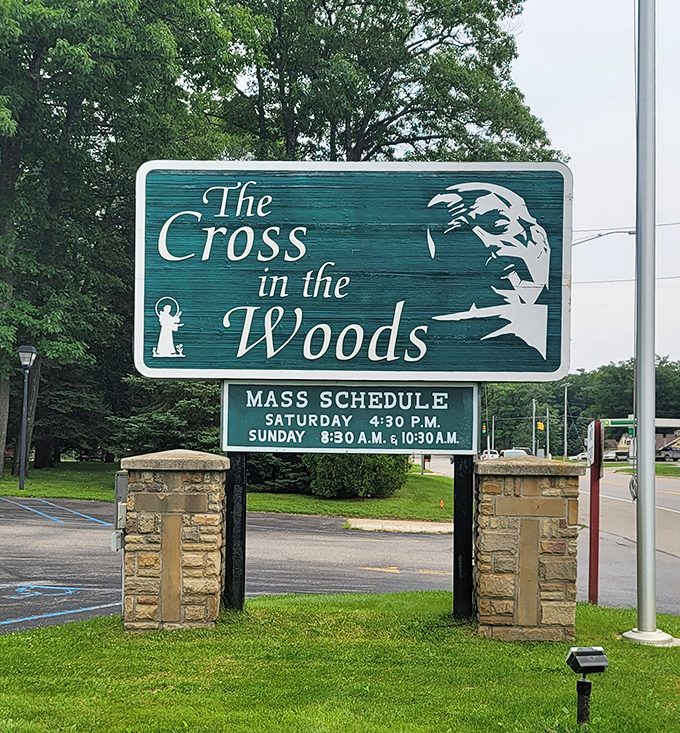 The welcoming sign for Cross in the Woods announces mass times, inviting visitors to experience this unique spiritual sanctuary in Michigan's north woods.