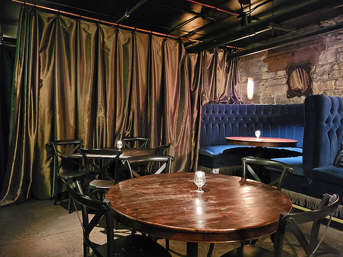 Dining Tables: Rich wooden tables surrounded by tufted seating create private islands for conversation beneath dramatically draped fabric and exposed ductwork.