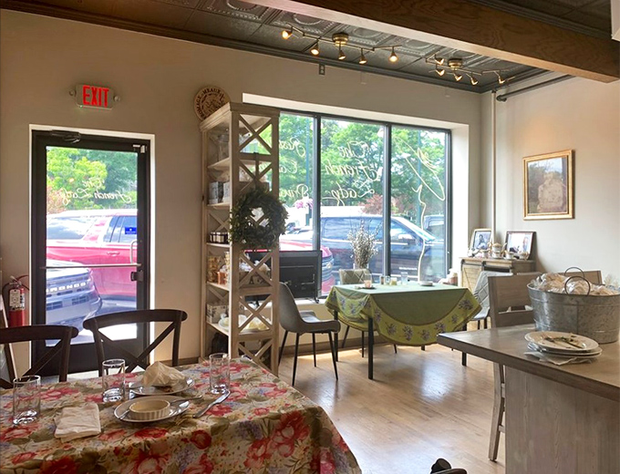 Sunlight streams through large windows, illuminating the cozy interior where floral tablecloths and simple elegance create a genuine French ambiance.