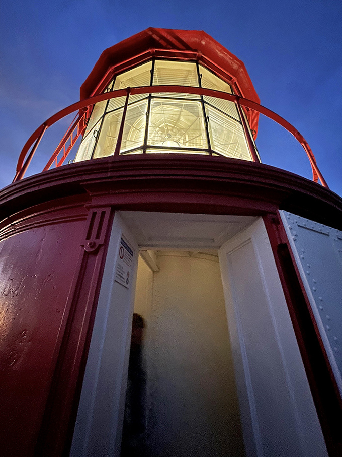 The lighthouse's lantern room glows crimson against twilight blue, housing the original Fresnel lens that still guides ships safely home.