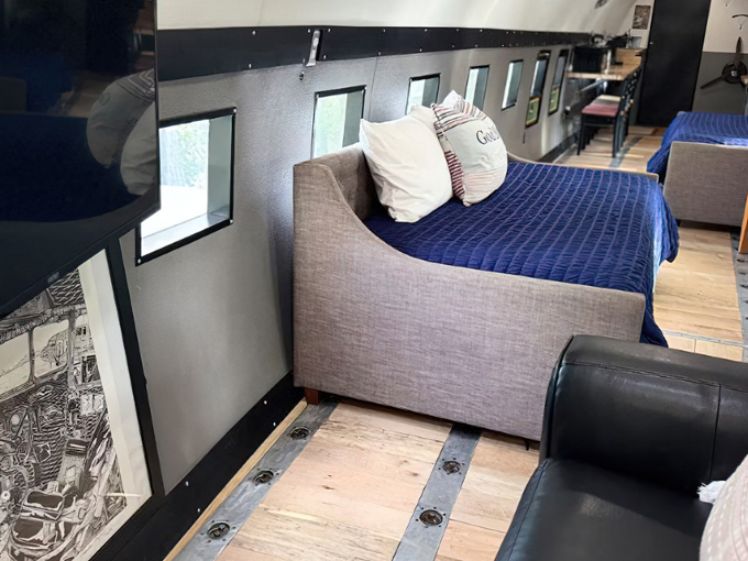 The aircraft's interior features a comfortable bed with blue bedding, surrounded by windows framing forest views.