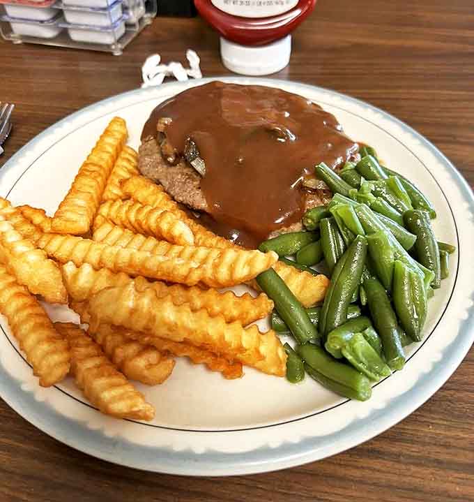 This hamburger steak smothered in rich gravy alongside crispy fries and fresh green beans is the definition of comfort on a plate.