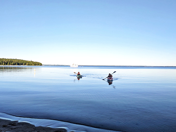 Kayakers glide across mirror-like waters, their paddles creating ripples in the perfect reflection of Michigan's blue skies.