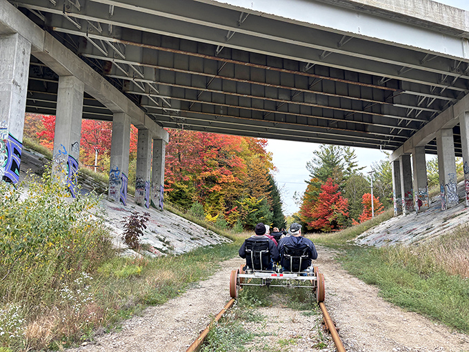 Nature frames the journey perfectly as rail bikers pass beneath a highway overpass, where autumn colors create a stunning natural tunnel.