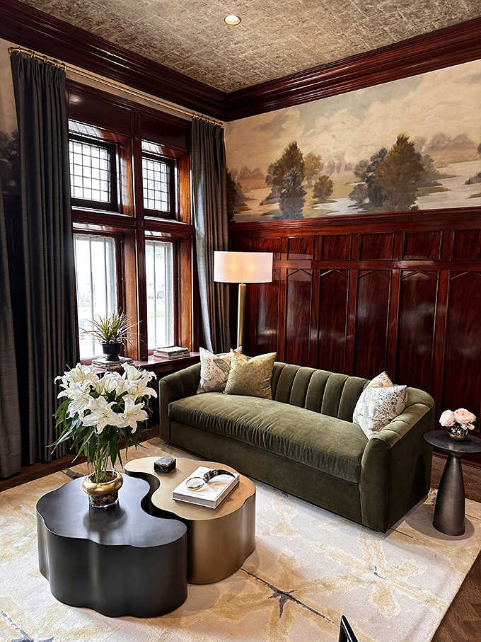 Rich wood paneling and a velvet sofa create a sanctuary for conversation, where painted landscapes above seem to extend the room into nature.