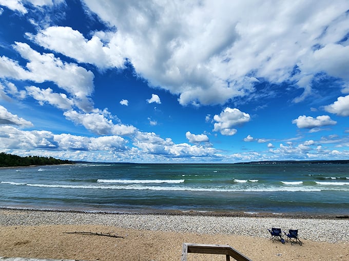 The beach at Petoskey State Park stretches invitingly beneath dramatic skies, where two empty chairs await someone wise enough to pause.