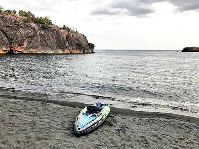A lone kayak rests on the distinctive dark sand, ready for exploration of Lake Superior's hidden coves.