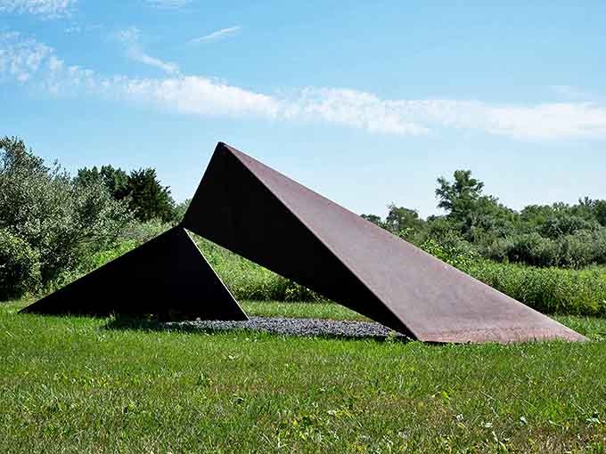 Rust-colored triangles slice through space with mathematical precision, creating shadows that move across the grass like sundials marking time.