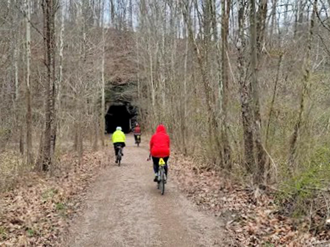 Bikers: Colorful cyclists approach the dark mouth of history, their modern gear contrasting with the tunnel's ancient, weathered embrace.