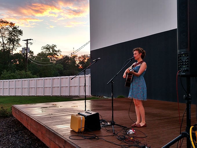 A performer strums her guitar against a painted sunset backdrop &ndash; live music before the movie adds another layer to the drive-in experience.