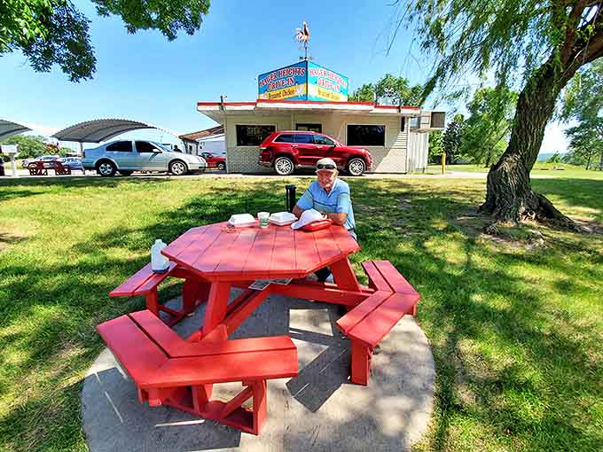 A lone diner savors the peaceful ambiance at a cherry-red picnic table, proving that sometimes the best company is excellent food.